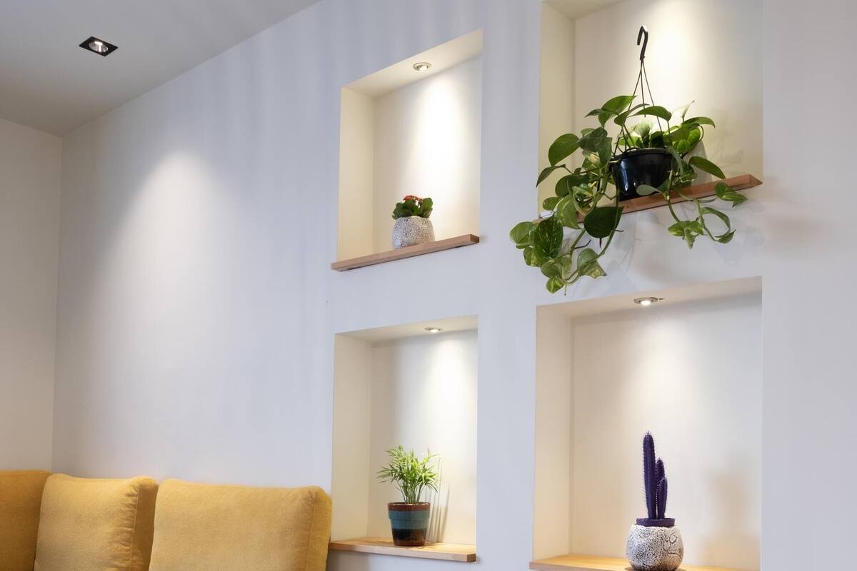 Uma sala de estar moderna com um sofá, uma mesa de centro de madeira com um vaso de flores e prateleiras fixadas na parede com plantas e objetos decorativos.