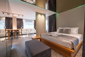 Quarto de hotel moderno com uma cama grande, área de estar com mesa e cadeiras, e vista através de um painel de vidro.