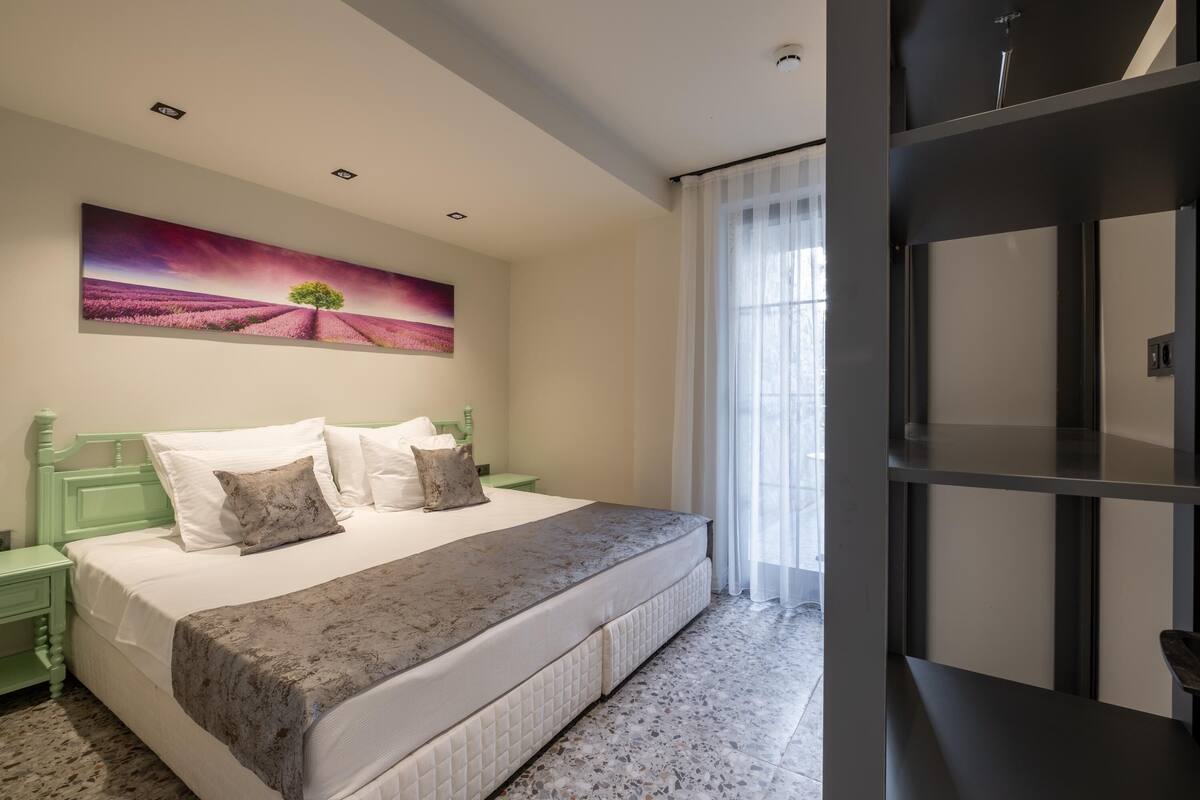 Quarto moderno com uma cama grande, uma mesa de cabeceira verde, um quadro de um campo com uma árvore e uma janela ampla com cortinas leves.