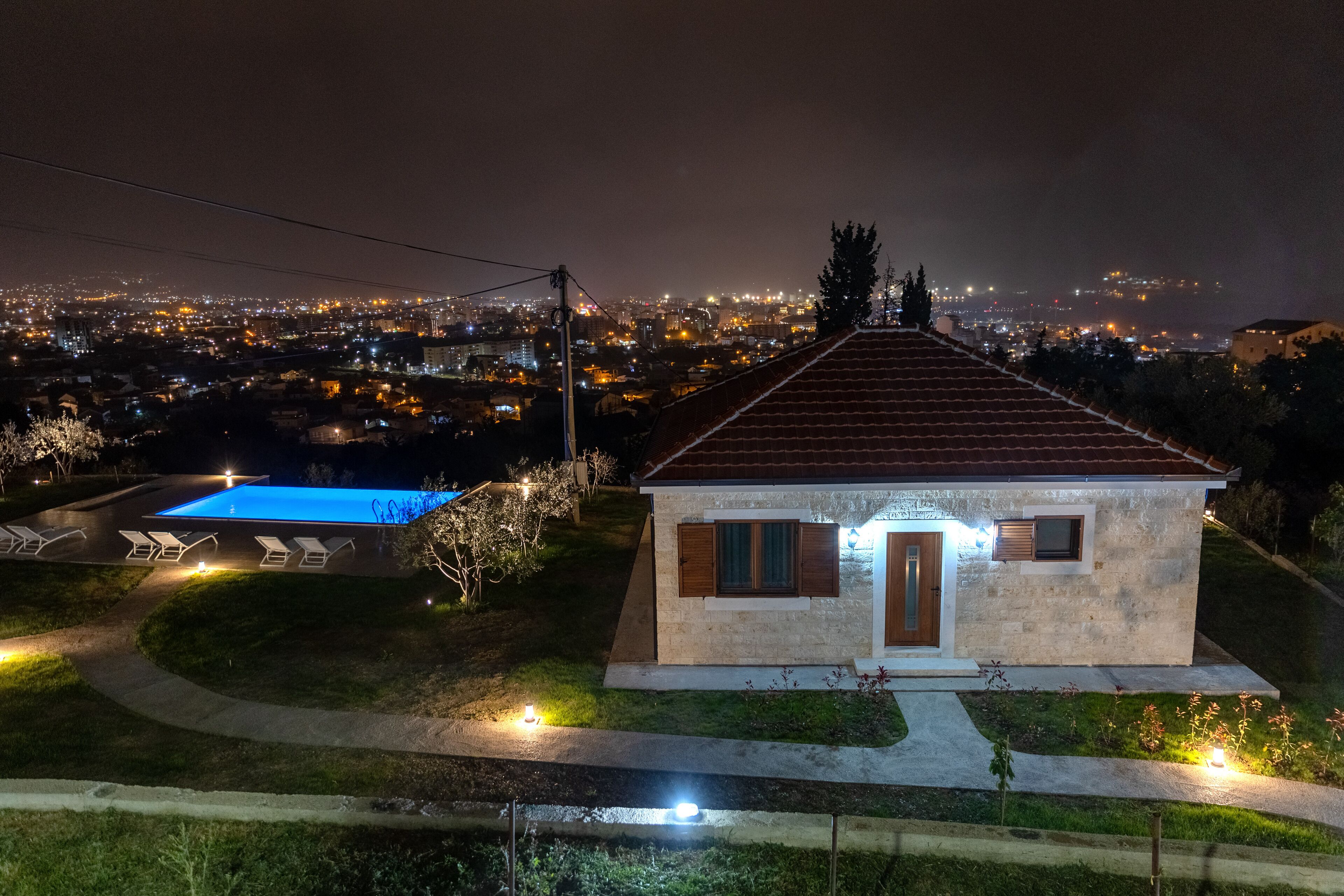 Uma casa com piscina, um pequeno jardim e vista para a paisagem urbana à noite.