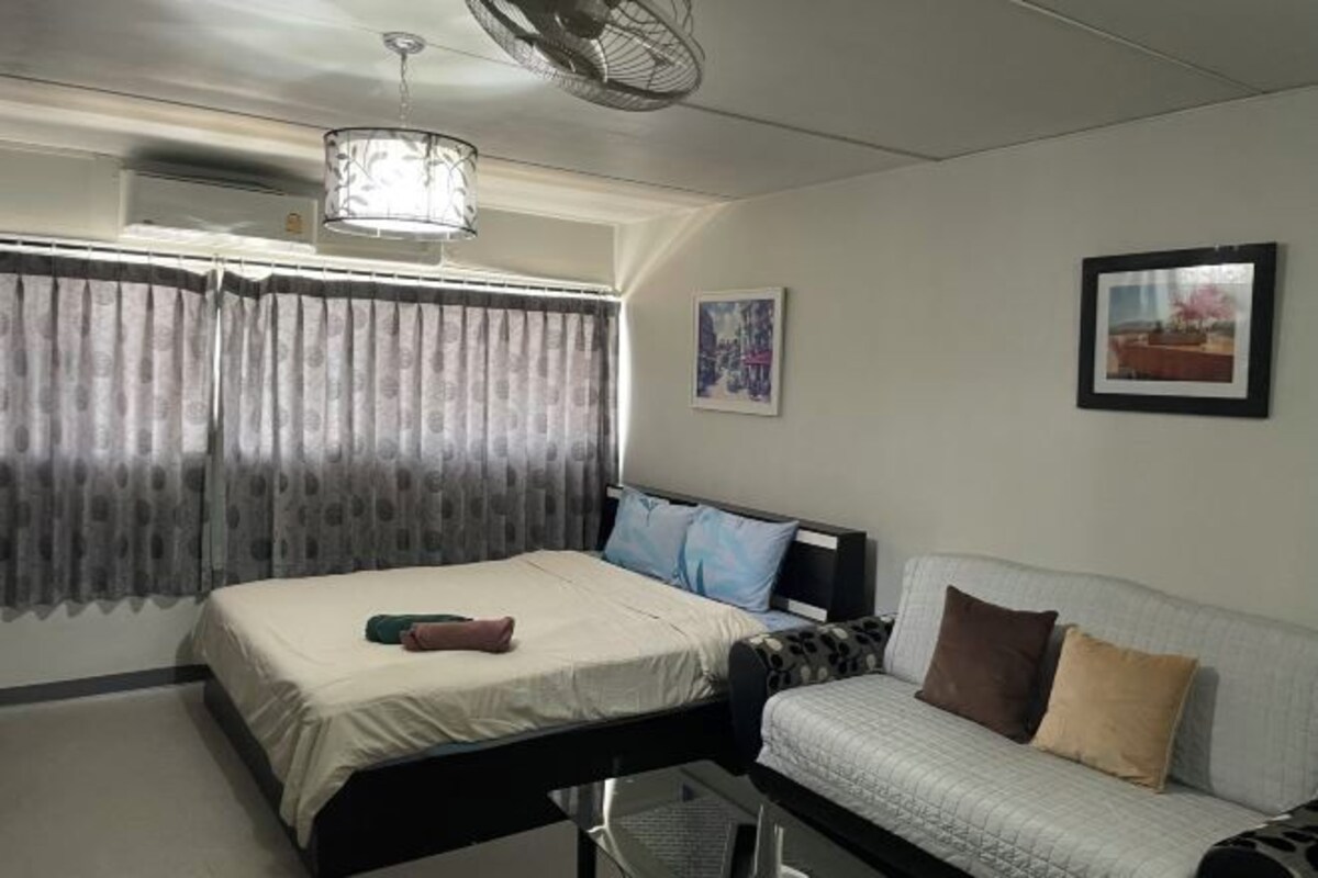 Quarto de hotel com uma cama, um sofá, um ventilador de teto e duas fotografias emolduradas na parede.