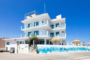 Um hotel moderno com fachada branca e detalhes em azul, apresentando uma escadaria que leva à entrada, um mural com vida marinha e um céu azul límpido.