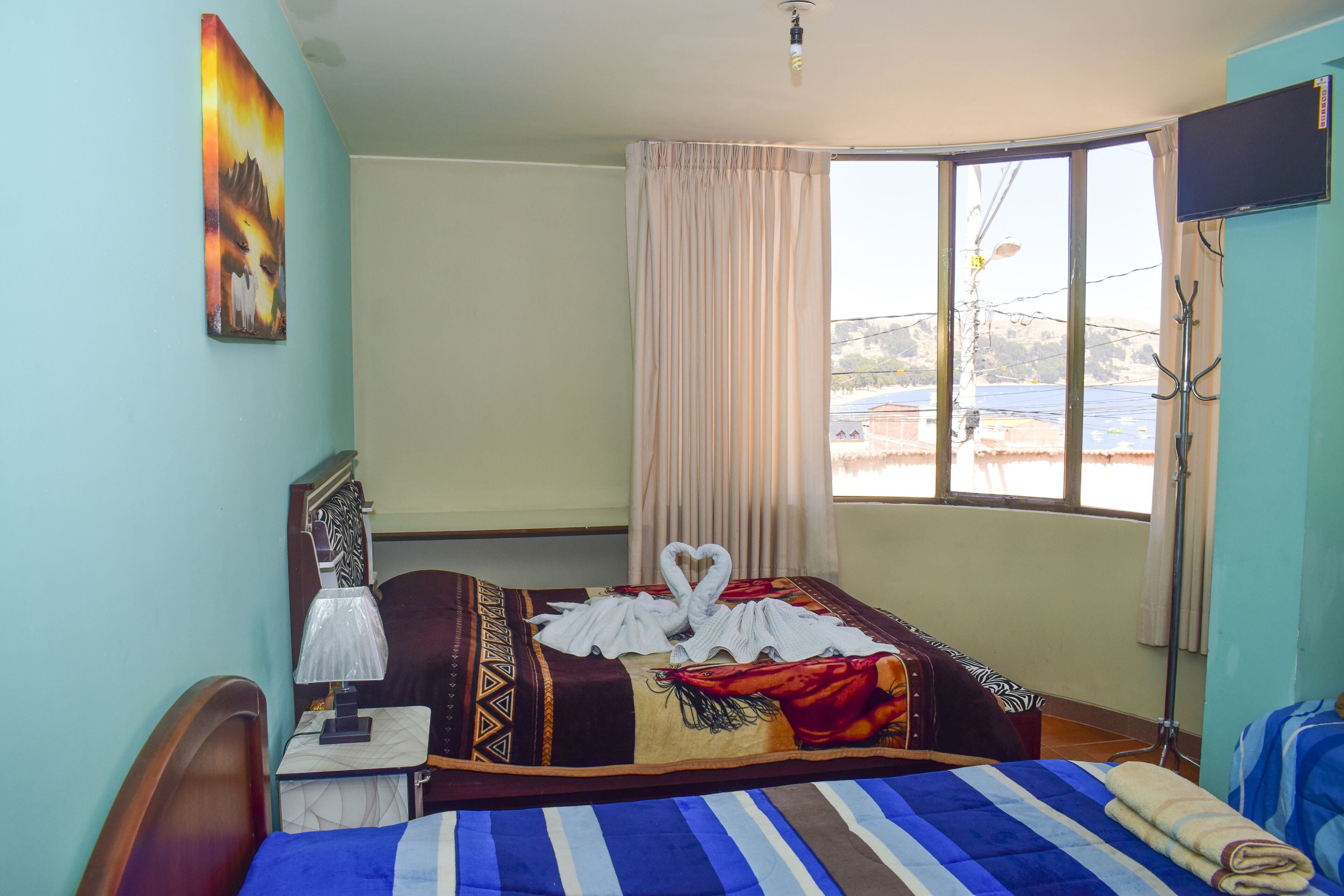 Quarto de hotel com uma cama, mesa de cabeceira com abajur, janela com cortinas e um quadro na parede.