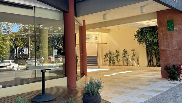 Entrada de hotel moderna com fachada de vidro, área de estar externa e plantas em vasos.