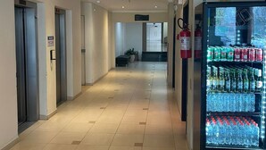 Corredor de hotel com pisos ladrilhados, elevadores, máquina de vendas automática e extintor de incêndio.
