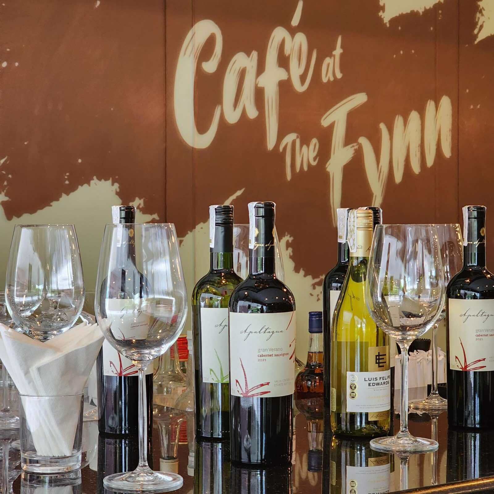 Uma área de bar com garrafas de vinho e copos, com uma placa ao fundo que diz "Cafe at The Fynn".