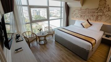 Quarto de hotel com uma cama grande, televisão, uma mesinha com cadeira e vista da cidade através de grandes janelas.