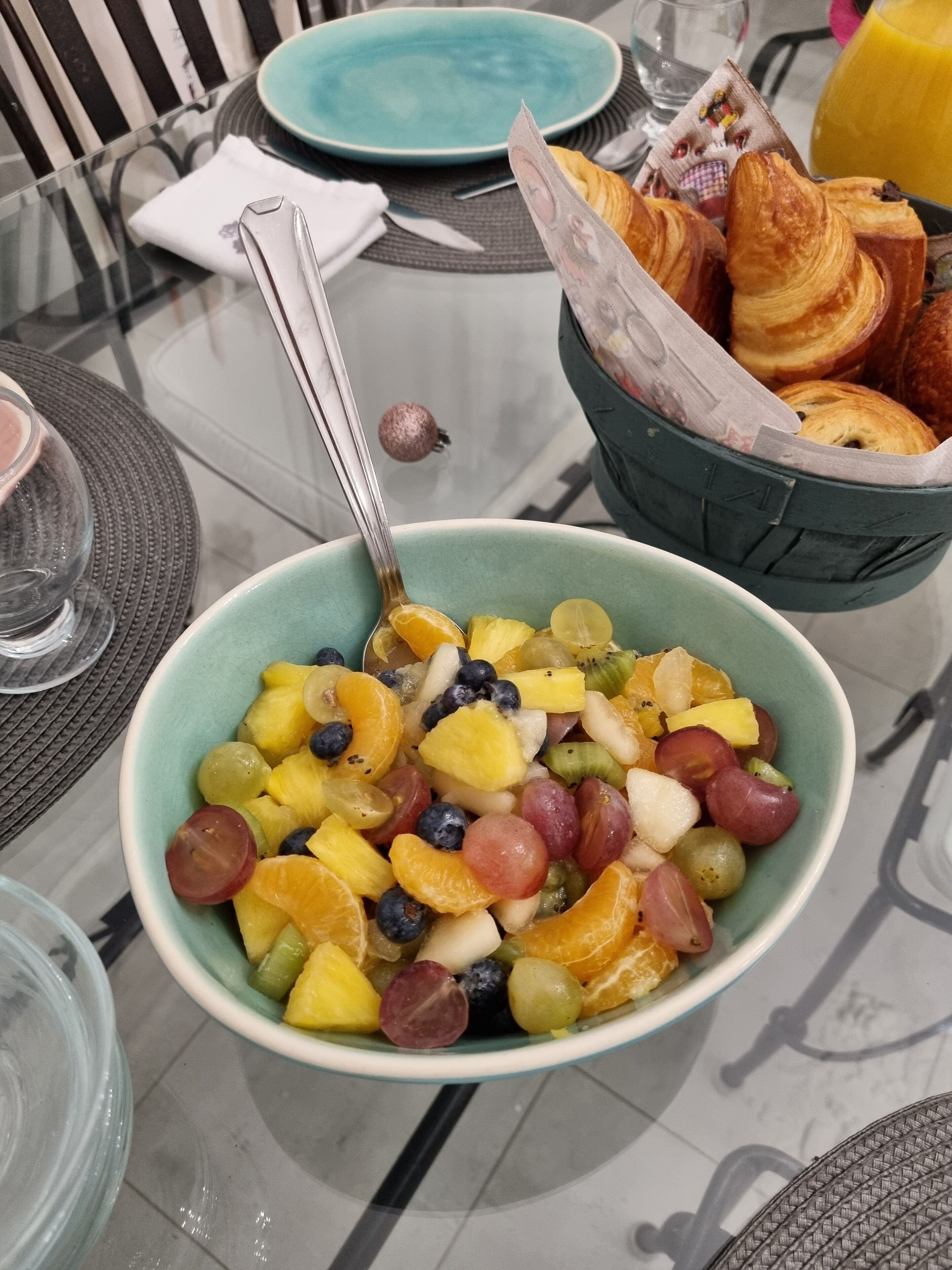 Disposição para o pequeno-almoço com uma tigela de salada de frutas, uma cesta de croissants e um copo de sumo de laranja sobre uma mesa.