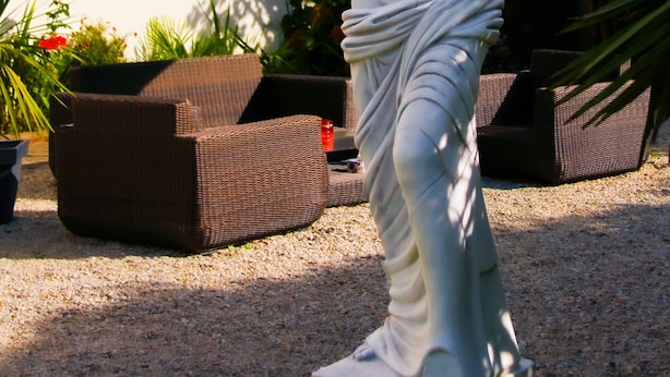 Estátua de uma mulher em um jardim, com guarda-chuva azul, móveis de vime e um edifício ao fundo.