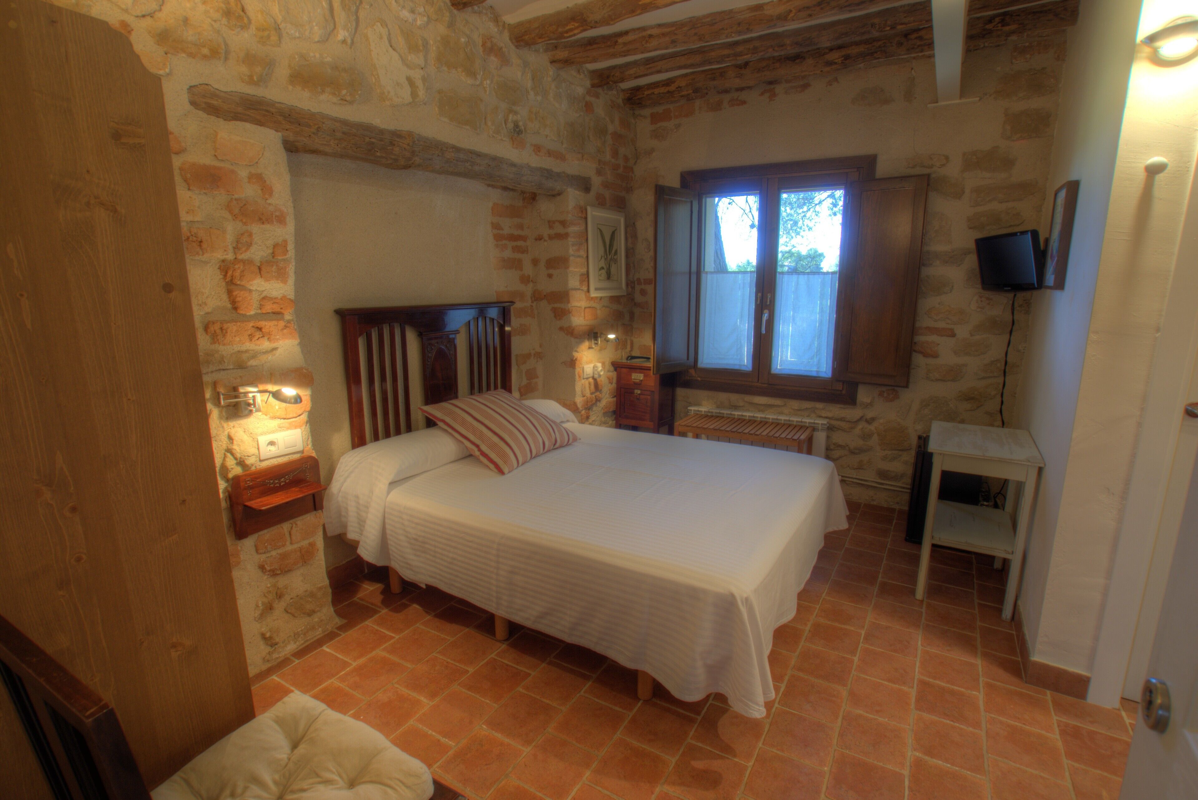 Quarto com cama, cabeceira de madeira, janela com cortinas, televisão fixada na parede e uma mesinha.