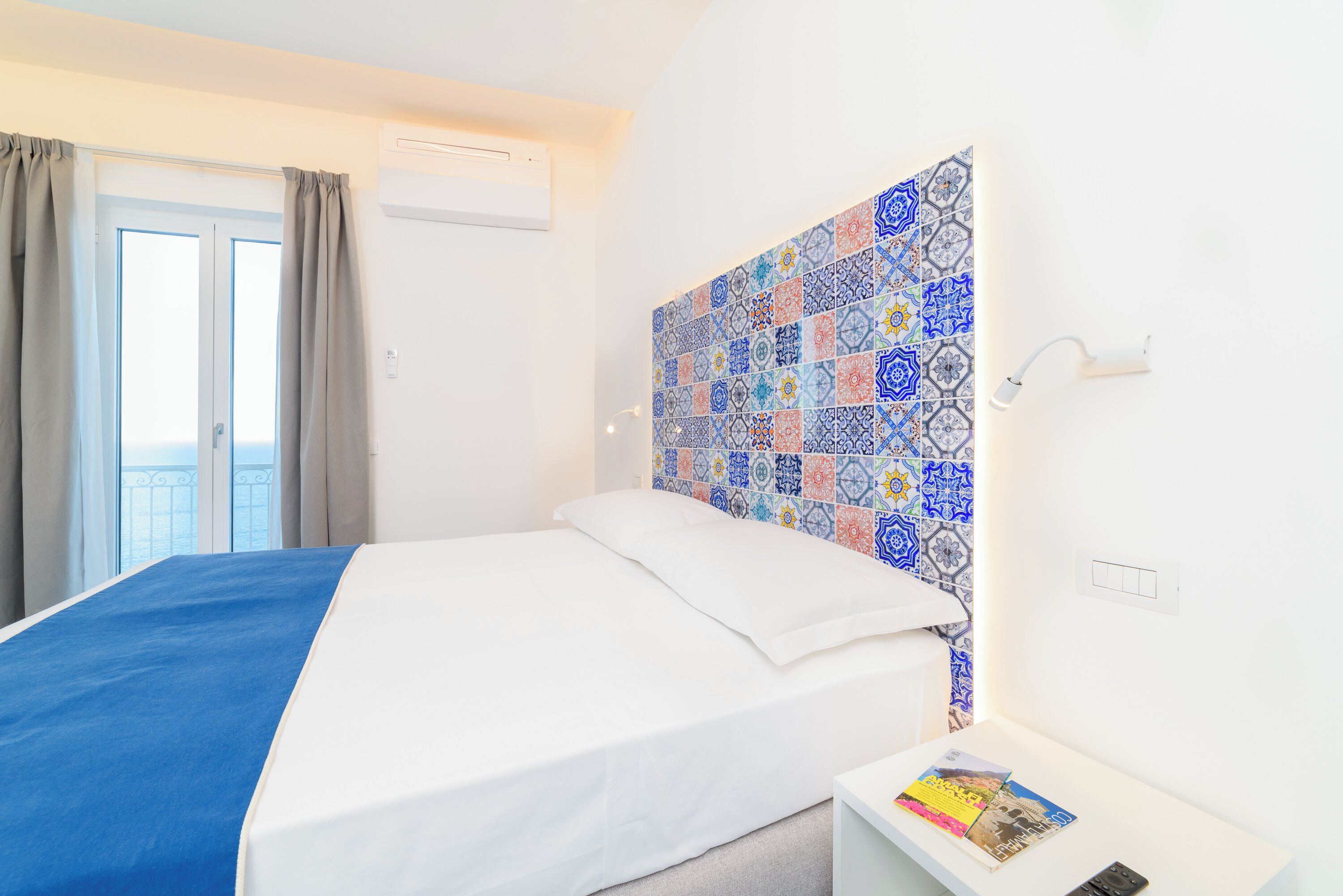 Quarto de hotel com cama, mesa de cabeceira, janela com cortinas e parede com design de azulejos estampados.