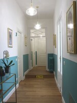 Um corredor com piso de madeira, paredes brancas e verde-água, e várias portas que dão para os quartos. Há quadros nas paredes e um vaso de planta em uma mesa.