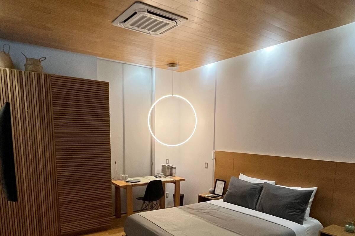Um quarto moderno com teto de madeira, cama com roupa de cama cinza, escrivaninha com cadeira e luminária redonda na parede.