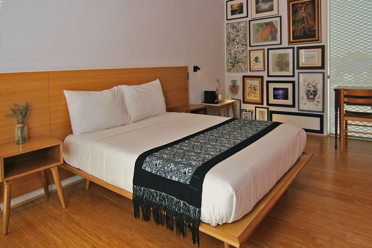 Cama bem arrumada com edredom branco e manta preta e branca, mesa de cabeceira de madeira com um vaso de plantas, uma cadeira de madeira e uma parede decorada com obras de arte emolduradas.
