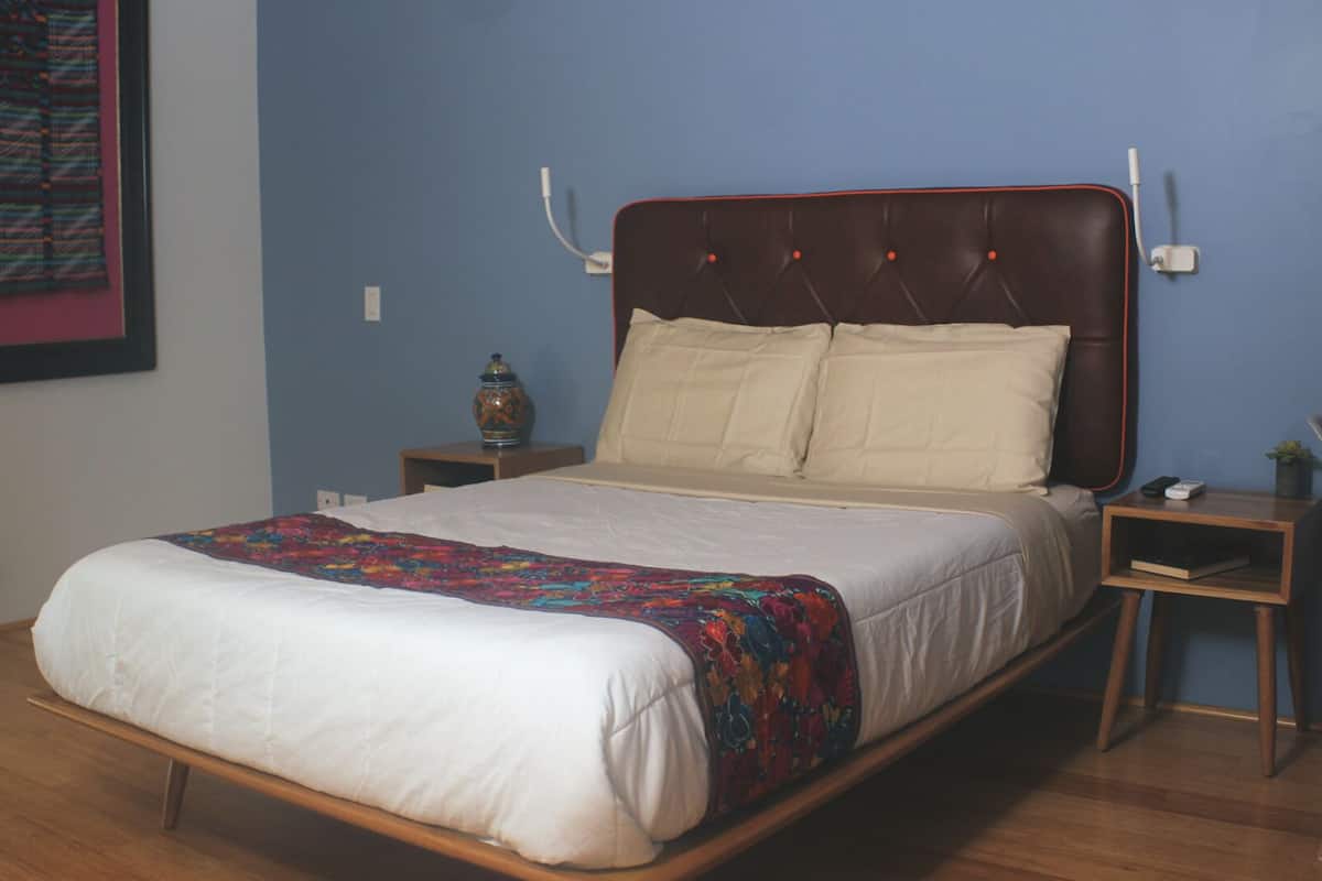 Uma cama bem arrumada com um edredom colorido, uma mesa de cabeceira com abajur e uma cadeira.