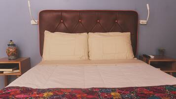 Cama com cabeceira estofada em tom de castanho, duas almofadas e um cobertor branco. Duas mesas de cabeceira, com um vaso e um controle remoto em uma delas.