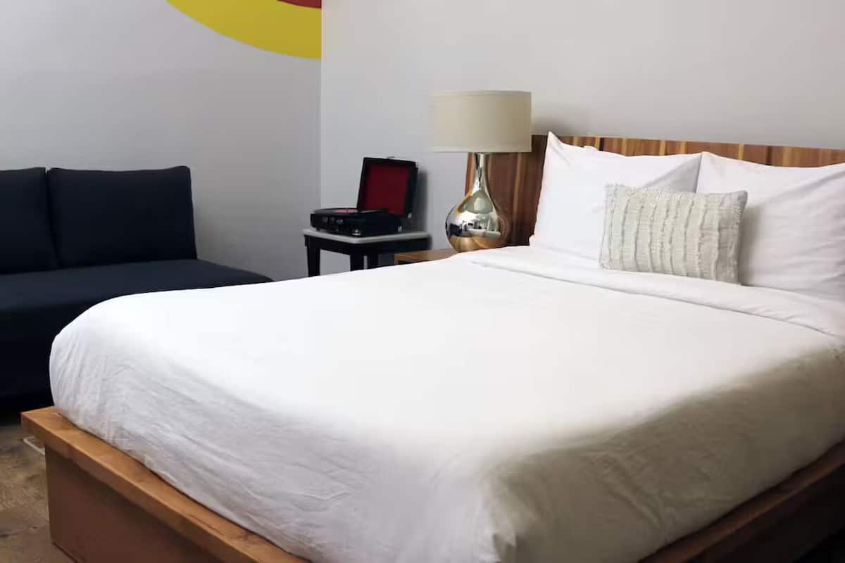 Quarto de hotel com uma cama, uma mesa de cabeceira com abajur, uma mesinha com uma mala vermelha e um sofá escuro.