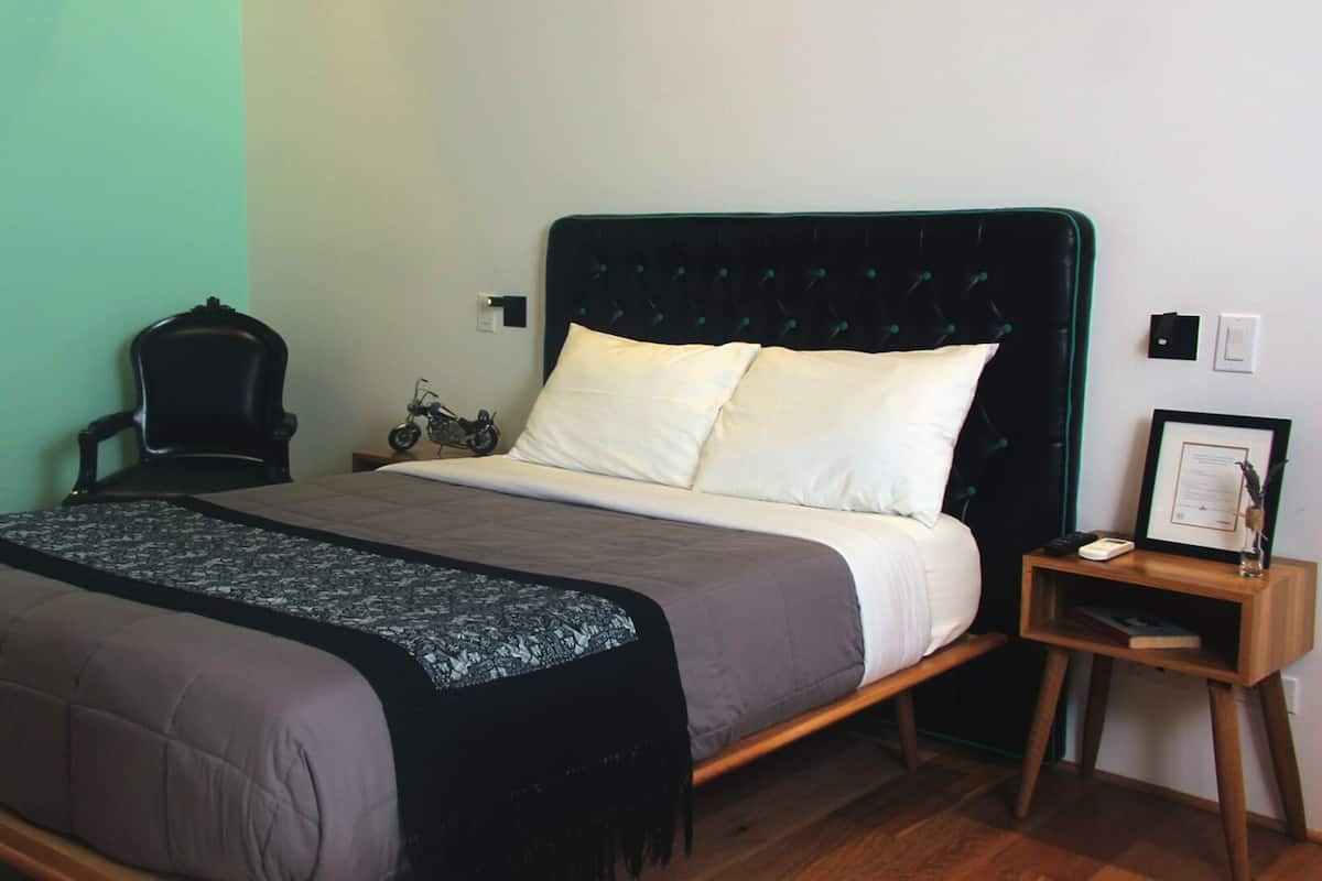 Cama bem arrumada com cabeceira escura, mesa de cabeceira com um certificado emoldurado e uma pequena luminária.