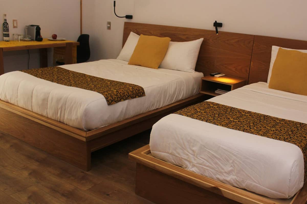 Duas camas de casal com estrutura de madeira e roupa de cama branca, uma pequena mesa de cabeceira com abajur, e uma escrivaninha de madeira com uma garrafa e copos.