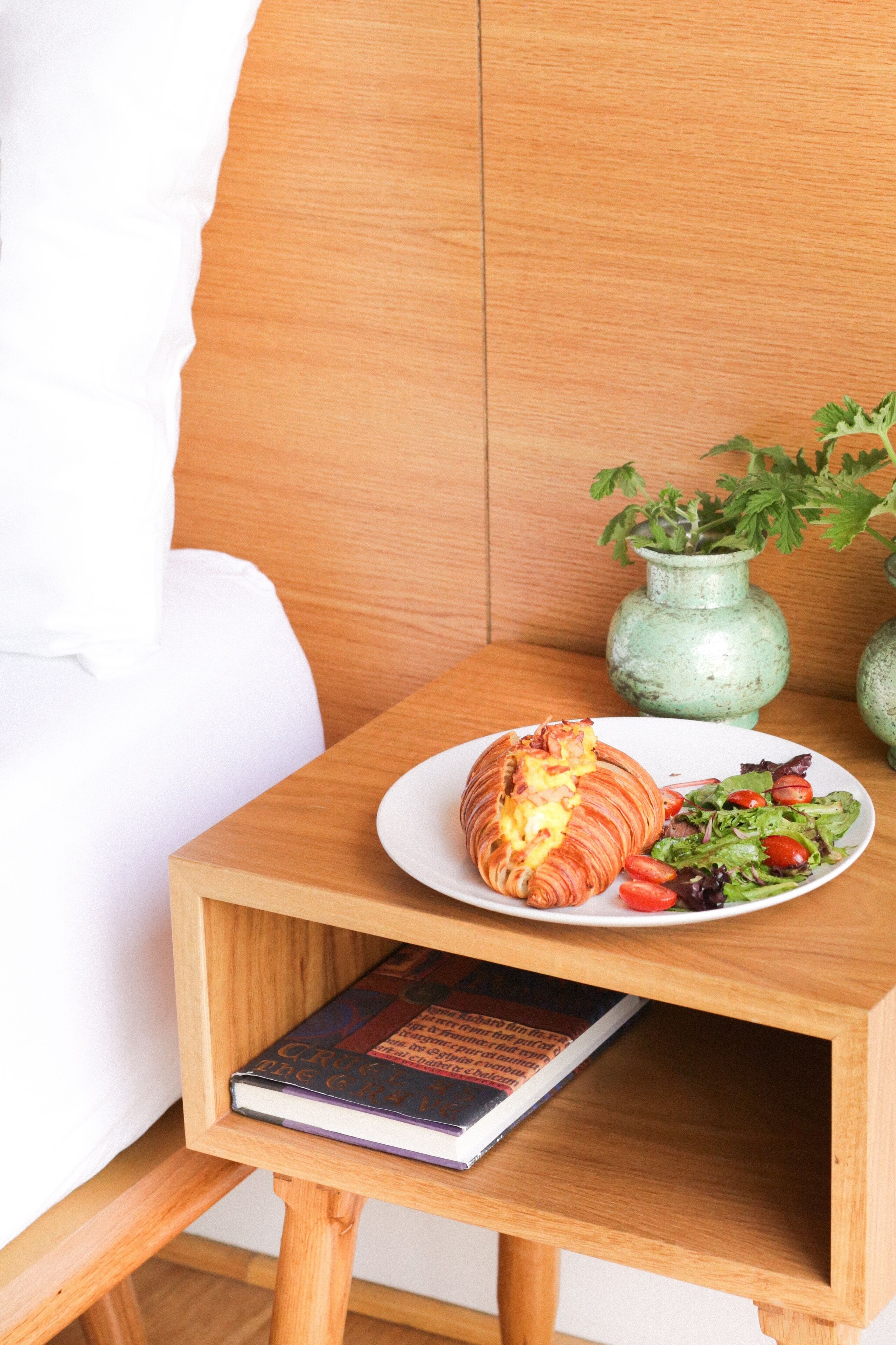 Uma mesa de cabeceira de madeira com um prato de comida, um livro e um vaso verde.