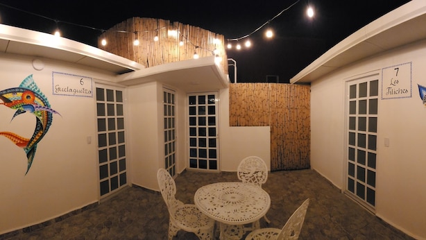 Um espaço coberto ao ar livre com paredes brancas, duas portas e uma mesa com cadeiras. Há luzes de corda acima e uma parede de bambu.