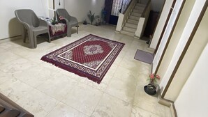 Uma sala de estar com um tapete vermelho estampado, duas cadeiras cinzas, uma mesinha, um vaso de planta e uma escada que dá para outro andar.