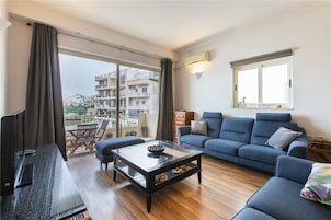 Uma sala de estar moderna com um sofá, uma mesa de centro de vidro, uma televisão e uma varanda com vista para edifícios.