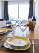 Uma mesa de jantar posta com pratos, tigelas, copos e talheres, com vista para uma sala de estar ao fundo.