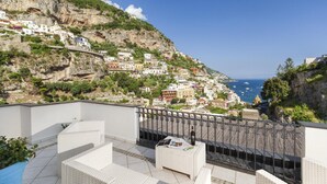 Terraço com mobiliário de vime, uma garrafa de vinho e vista para uma vila costeira com edifícios numa encosta.