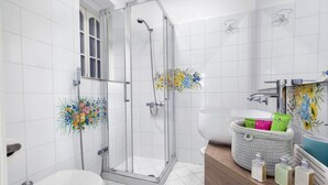 Uma casa de banho moderna com cabine de duche em vidro, sanita branca, lavatório com cesto decorativo e espelho com azulejos florais.