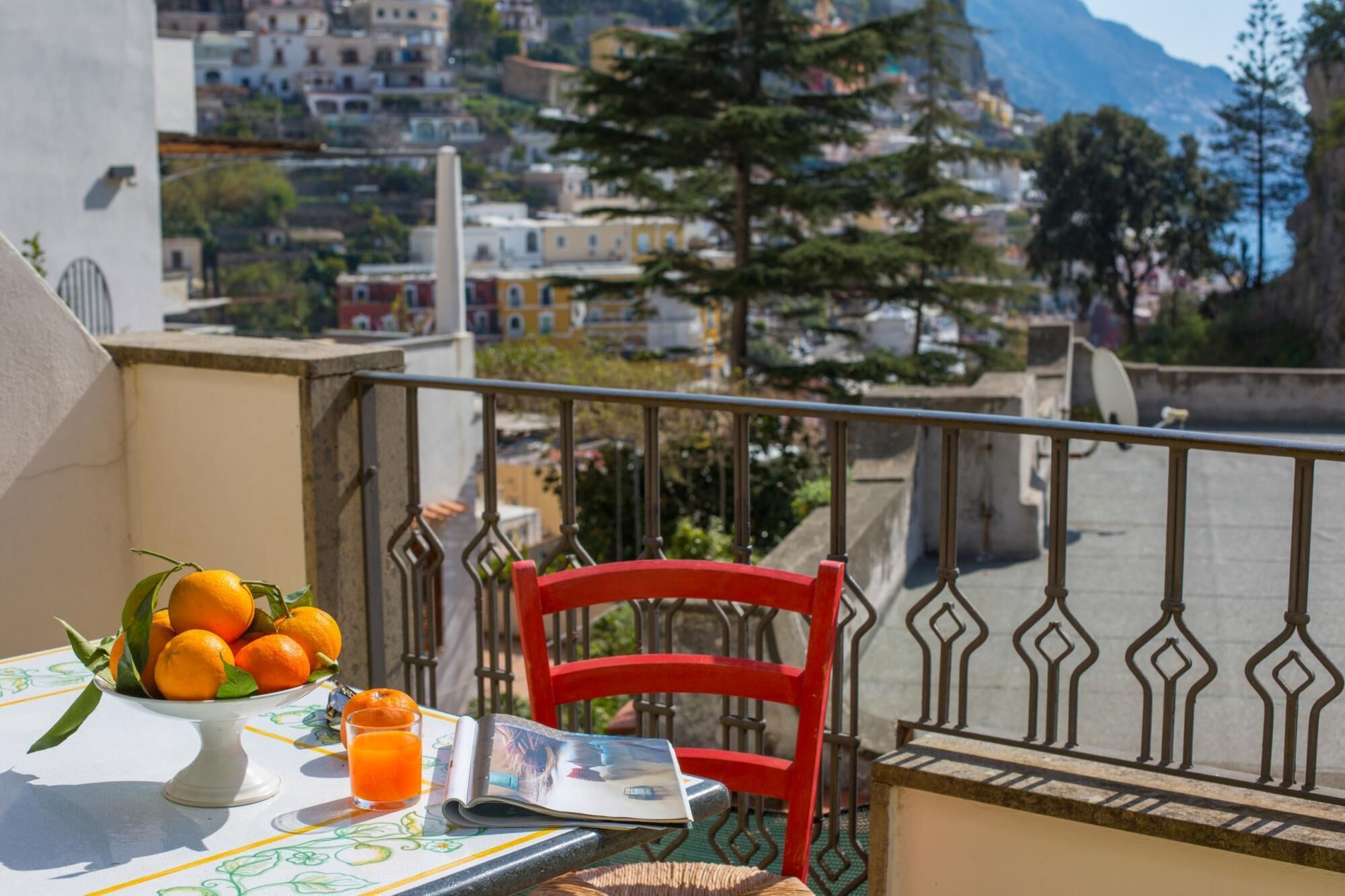 Uma varanda com uma mesa, uma cadeira vermelha, uma tigela de laranjas e um copo de suco de laranja.