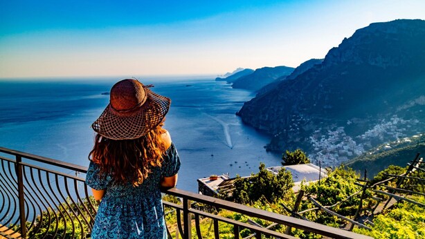 Mulher com vestido estampado e chapéu de abas largas observa uma paisagem costeira com vista clara para o mar e colinas ao longe.