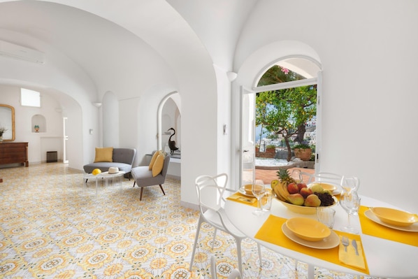 Uma sala de estar ampla e iluminada, com uma mesa de jantar posta para dois, um sofá e vista para o exterior.