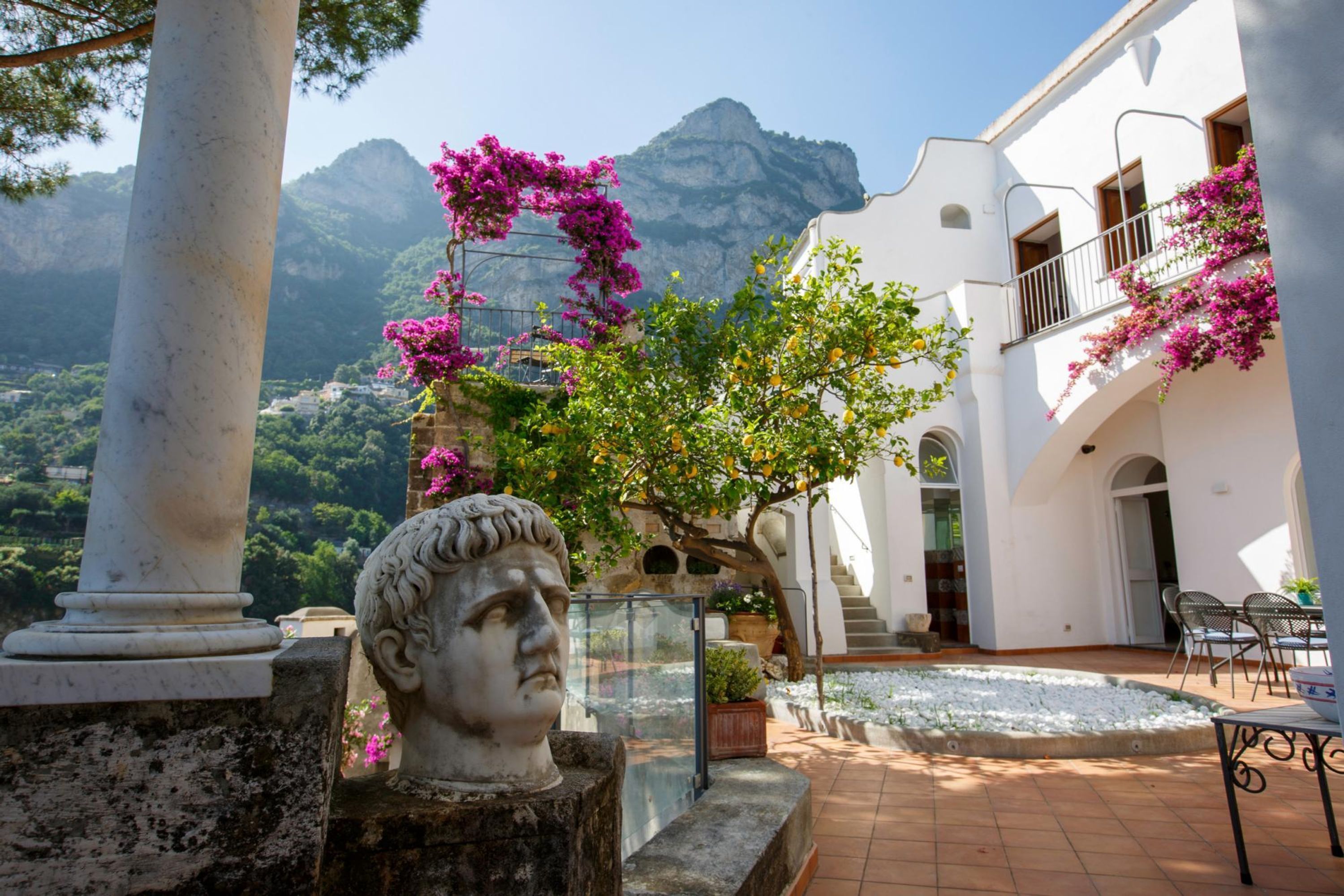Pátio com estátua clássica, edifício branco com varandas e paisagem montanhosa ao fundo.