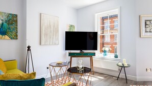 Uma sala de estar moderna com uma televisão de tela plana, um sofá amarelo, uma mesa de centro e vista para um edifício de tijolos por uma grande janela.