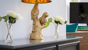 Um armário de madeira escura com uma luminária verde, dois vasos com flores brancas e uma figura decorativa de uma coruja.