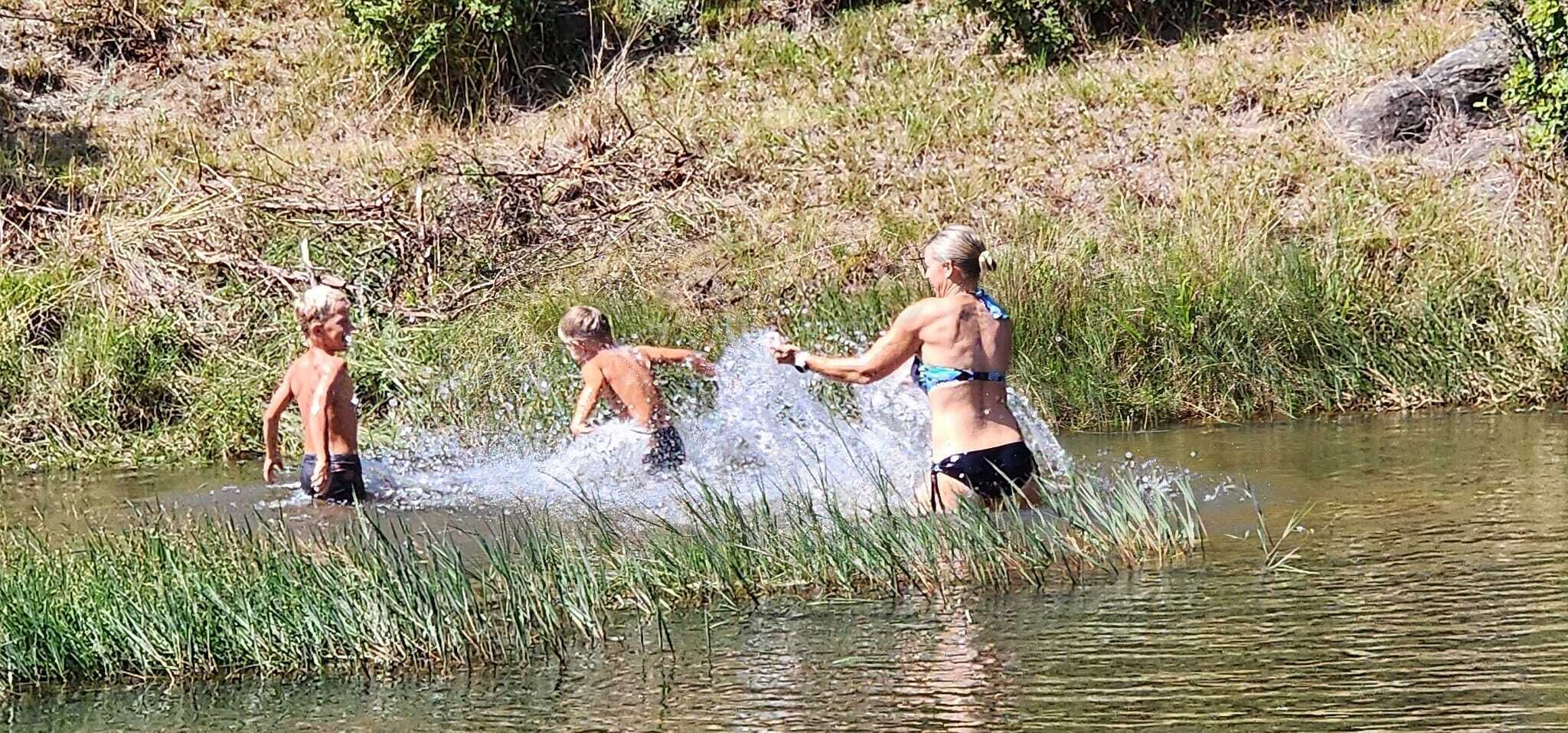 Três pessoas brincando em águas rasas, cercadas por grama e vegetação.