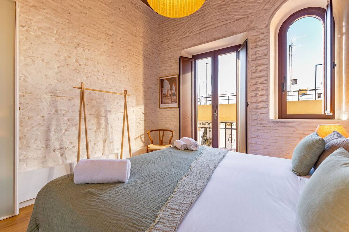 Quarto com parede de tijolos, cama com cobertor verde, cadeira de madeira e janela com vista para prédios.