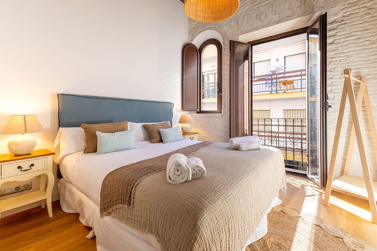 Quarto com uma cama grande, duas mesas de cabeceira com luminárias, piso de madeira, varanda com grade de ferro forjado e parede de pedra.