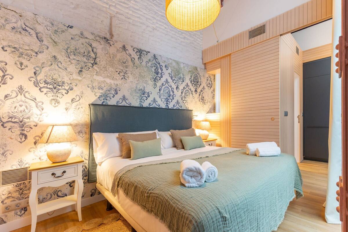 Cama bem arrumada com manta verde, mesa de cabeceira com abajur e papel de parede estampado.