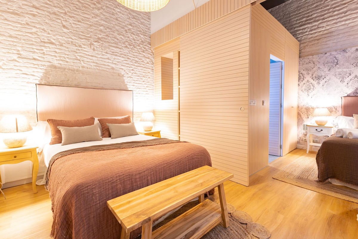 Quarto com uma cama grande, cabeceira de madeira, mesas de cabeceira com luminárias, um banquinho e uma parede texturizada.