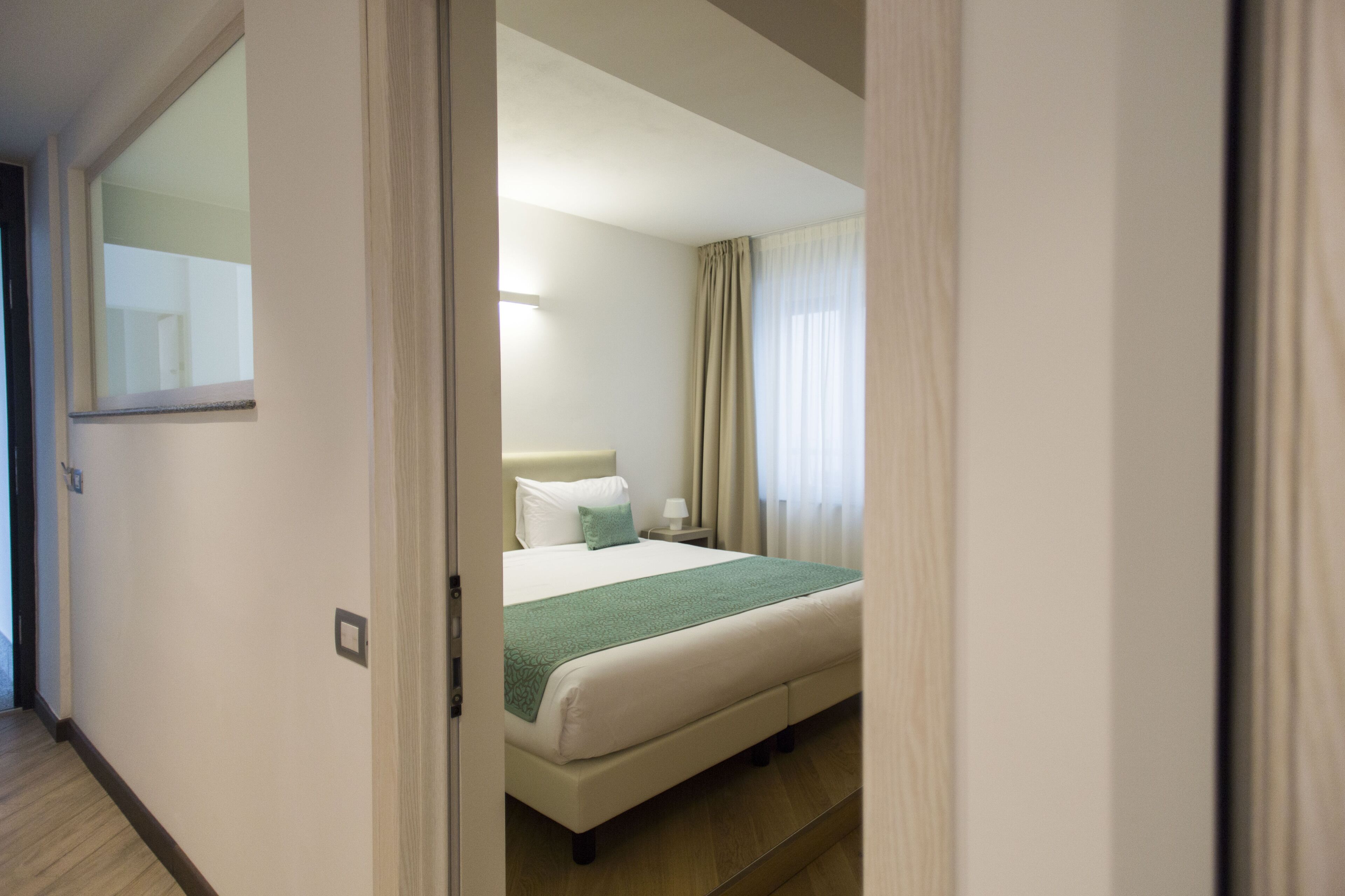 Quarto de hotel com cama, mesa de cabeceira, luminária e janela com cortinas.