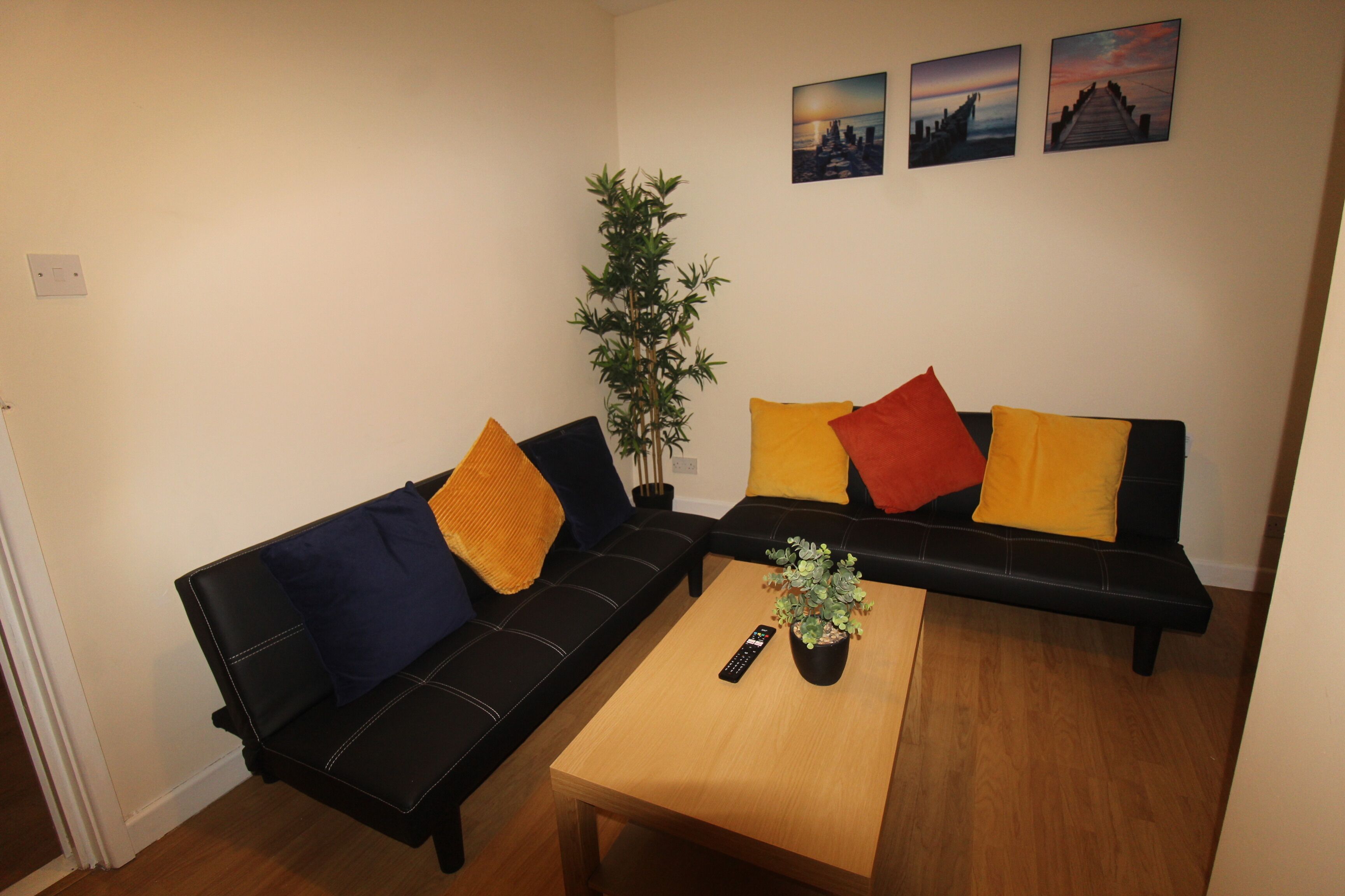 Uma sala de estar com um sofá preto, uma mesa de centro de madeira e um vaso de planta. Há três almofadas no sofá, e um controle remoto sobre a mesa.