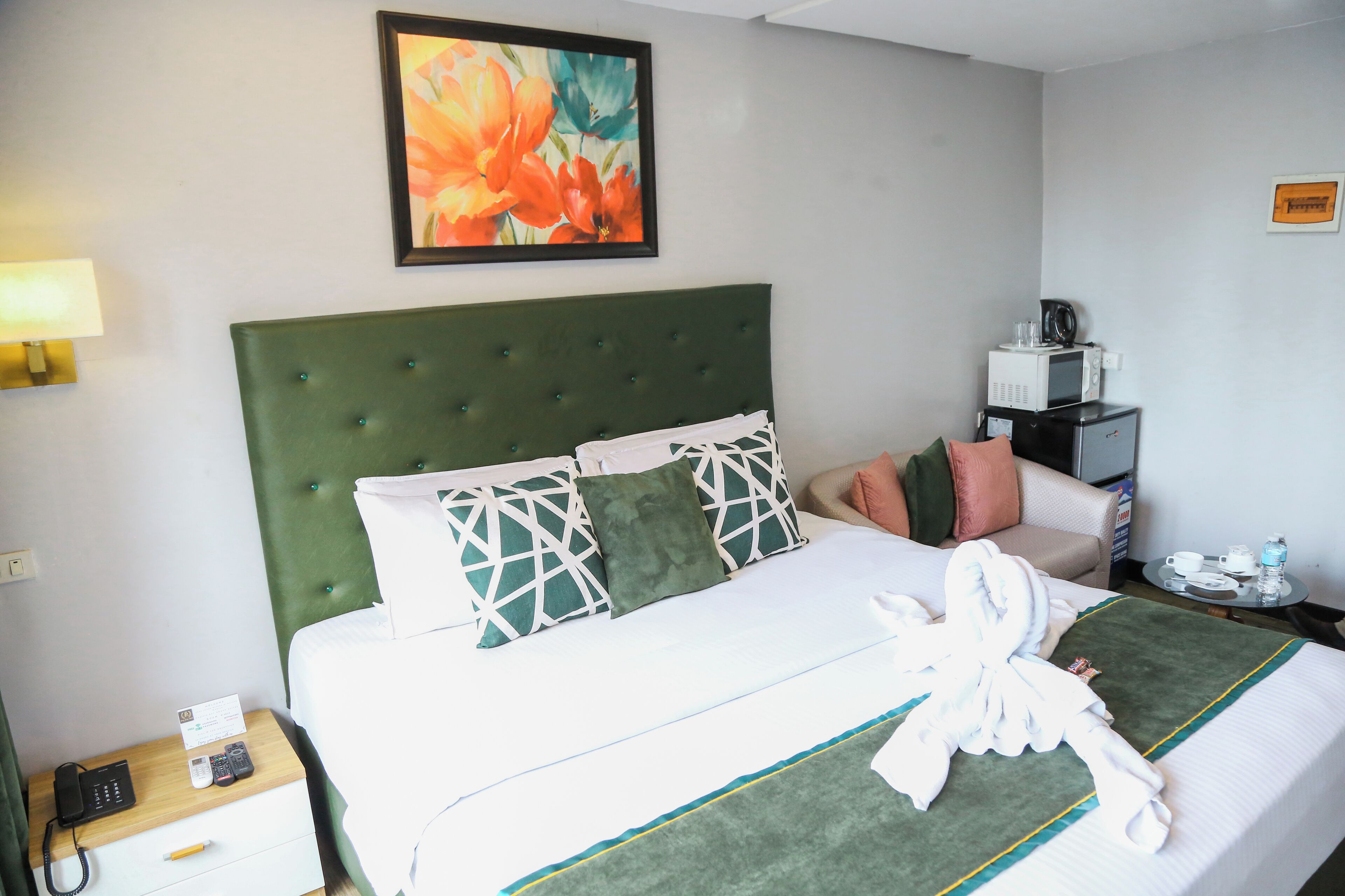 Quarto de hotel com cabeceira estofada em verde, cama com roupa de cama branca e verde, quadro de flores na parede, mesa de cabeceira com luminária, telefone e uma pequena bandeja com cafeteira, chávenas e garrafa de água.