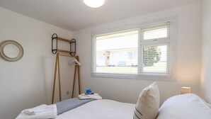 Cama bem arrumada com lençóis brancos, uma escada de madeira, um suporte pendurado e uma janela com persianas.