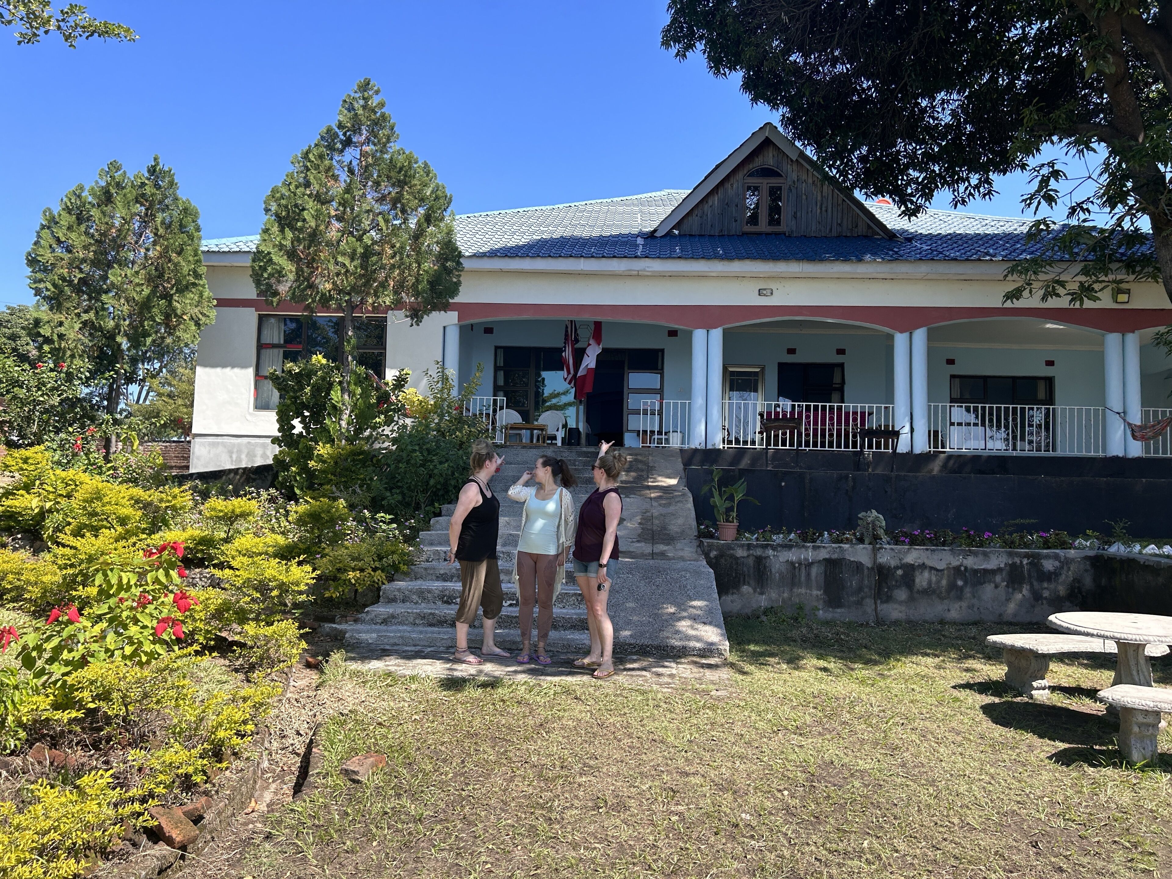 Um edifício com varanda coberta, degraus que levam à entrada e um jardim com plantas e um banco de pedra.