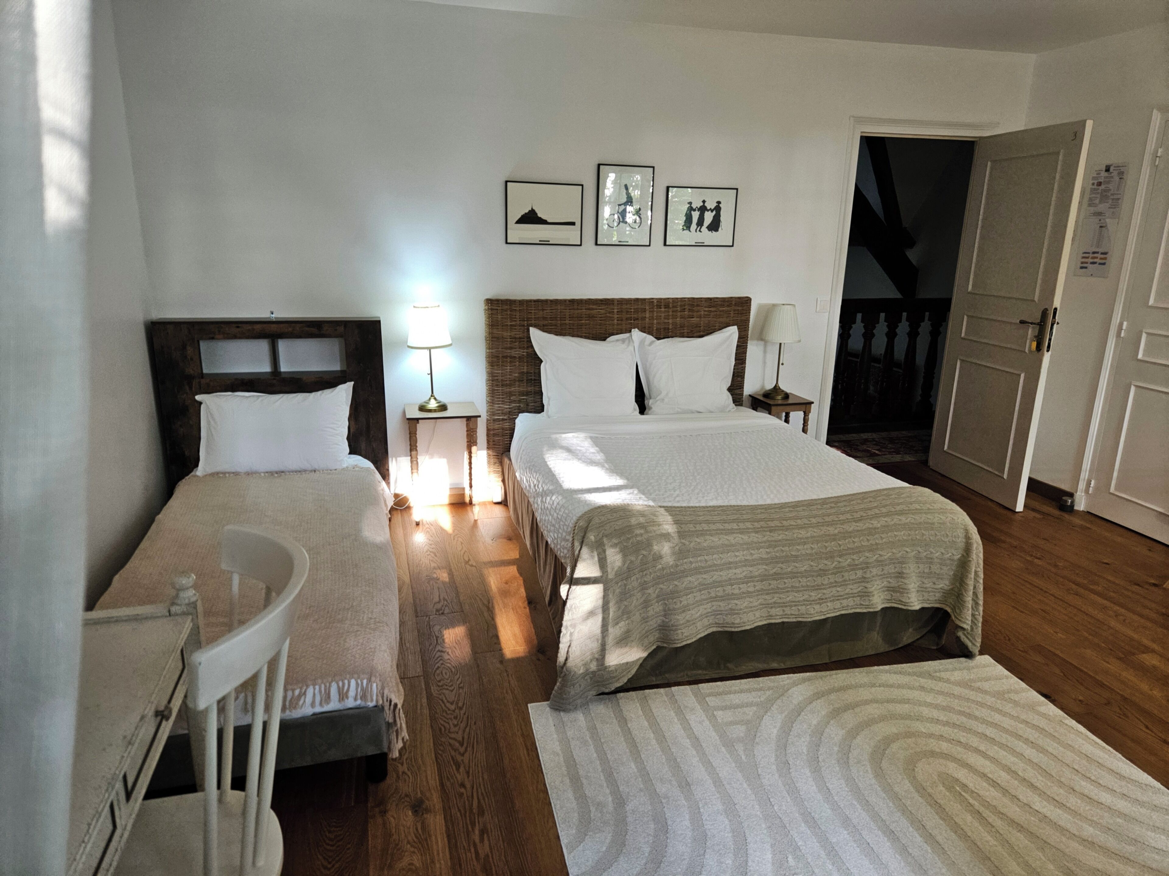 Quarto de hotel com duas camas, piso de madeira, uma cadeira branca e uma mesa pequena com luminárias. Há quadros nas paredes e uma porta à direita.
