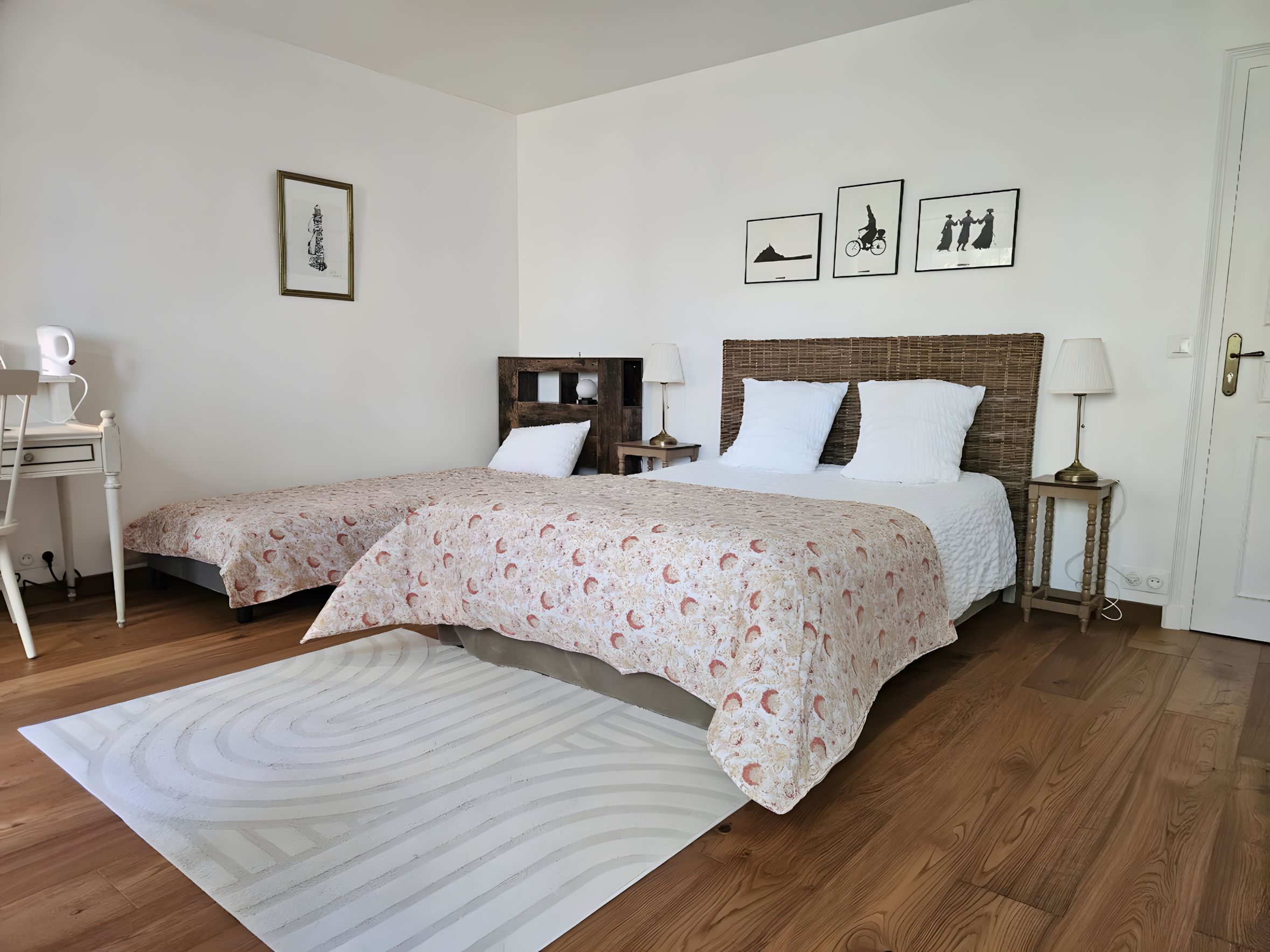 Quarto com duas camas, piso de madeira, tapete branco e parede com quadros emoldurados.