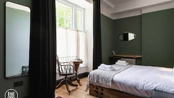 Quarto com cama grande, uma cadeira, mesa pequena, espelho e janela com cortinas.