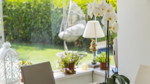 Uma mesa com um laptop, um vaso de planta e uma luminária. Há uma janela com vista para um jardim e um sebedo.